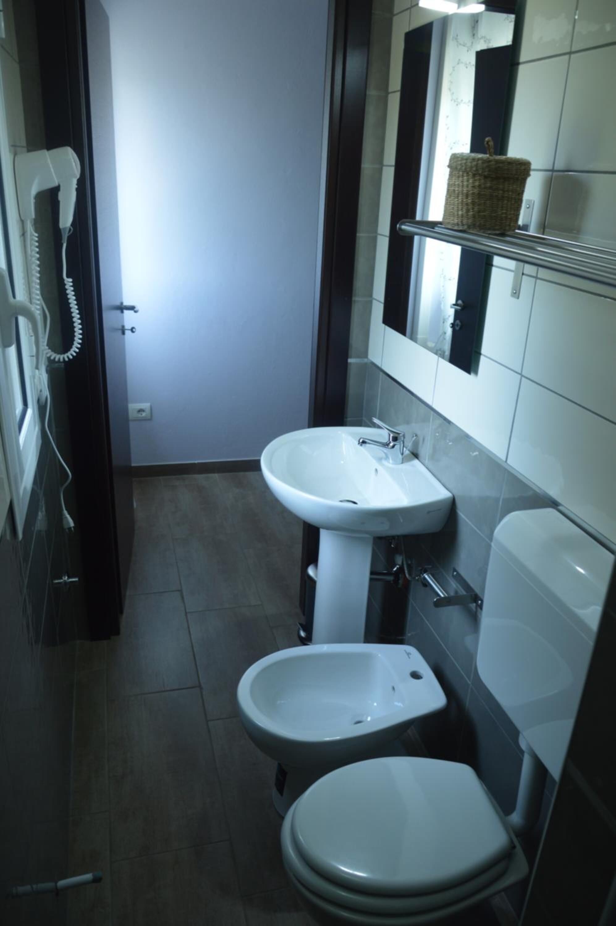 Casa de banho compacta com lavatório branco, sanita e bidé. Há um espelho sobre o lavatório e uma pequena prateleira com uma cesta.