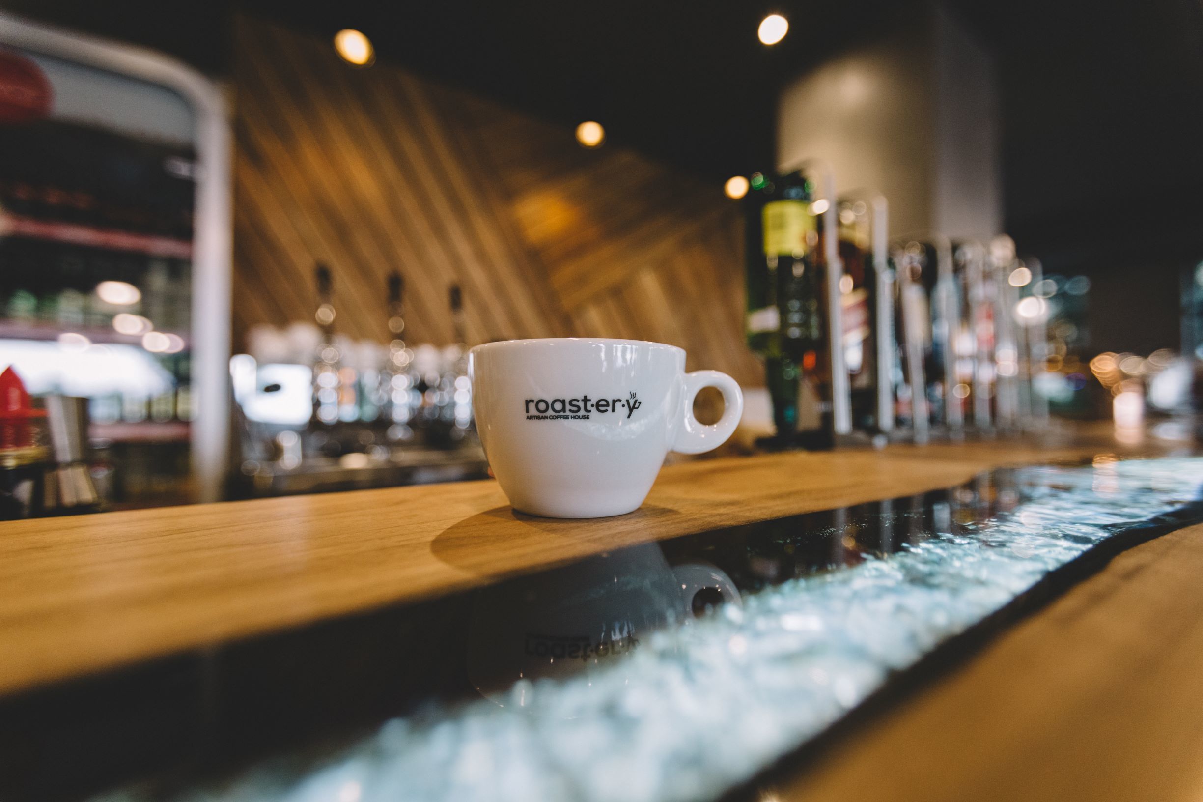 Uma chávena de café branca com o texto 'roastery' sobre um balcão de madeira.