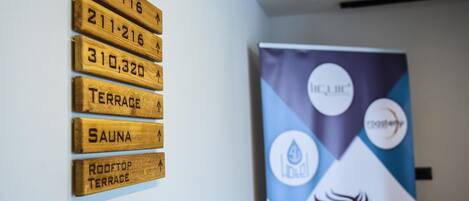 Placas de madeira com números e nomes de quartos, um cartaz com logotipos e uma saída de ventilação no teto.