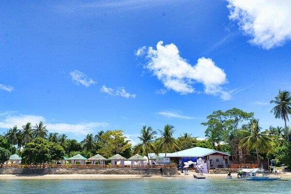 Resort à beira-mar com várias cabanas e um edifício grande, cercado por palmeiras e céu azul claro.