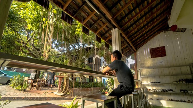 Um homem sentado em um banquinho sob uma área com telhado de madeira e plantas penduradas, em um ambiente rústico com parede de tijolos e exposição de sapatos.