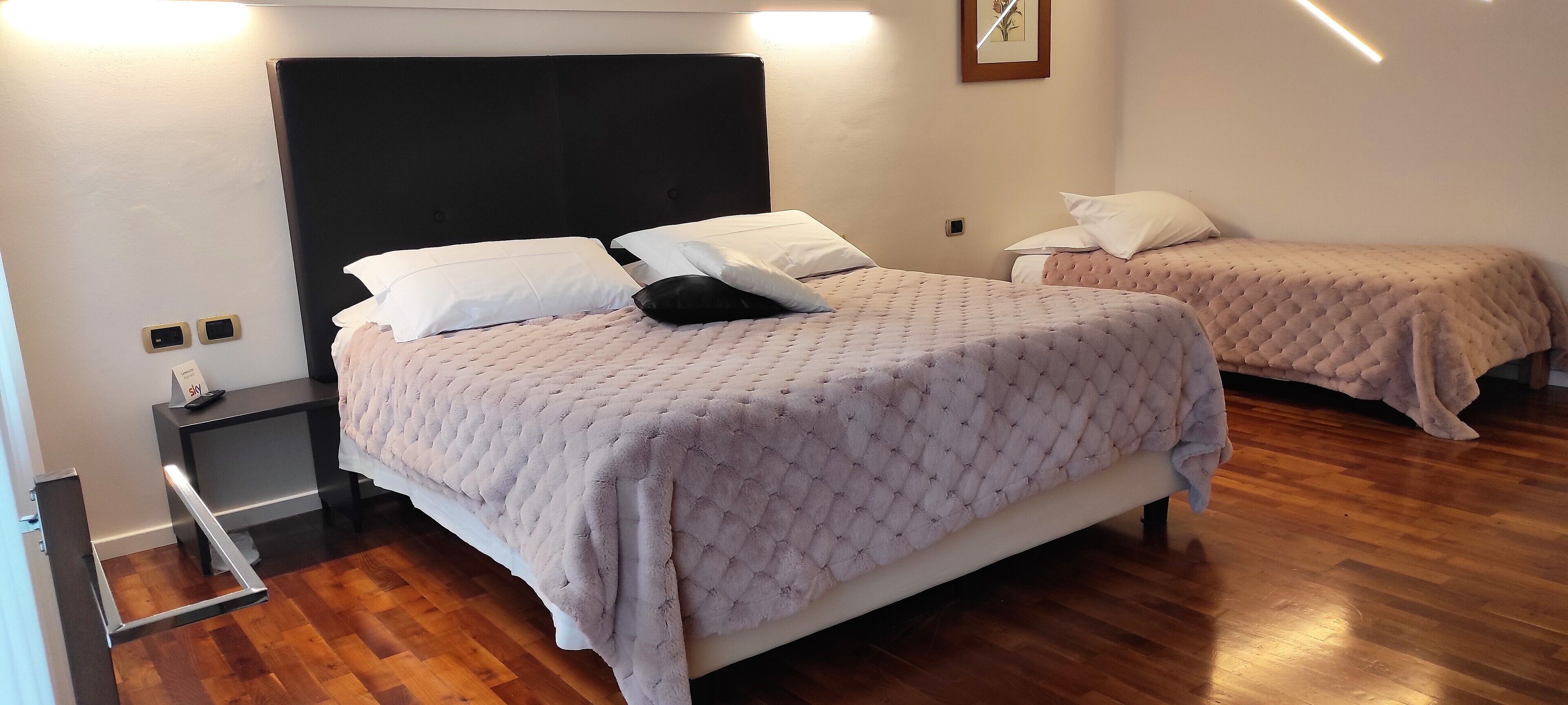 Quarto de hotel com duas camas, mesa de cabeceira, luminária na parede e piso de madeira.