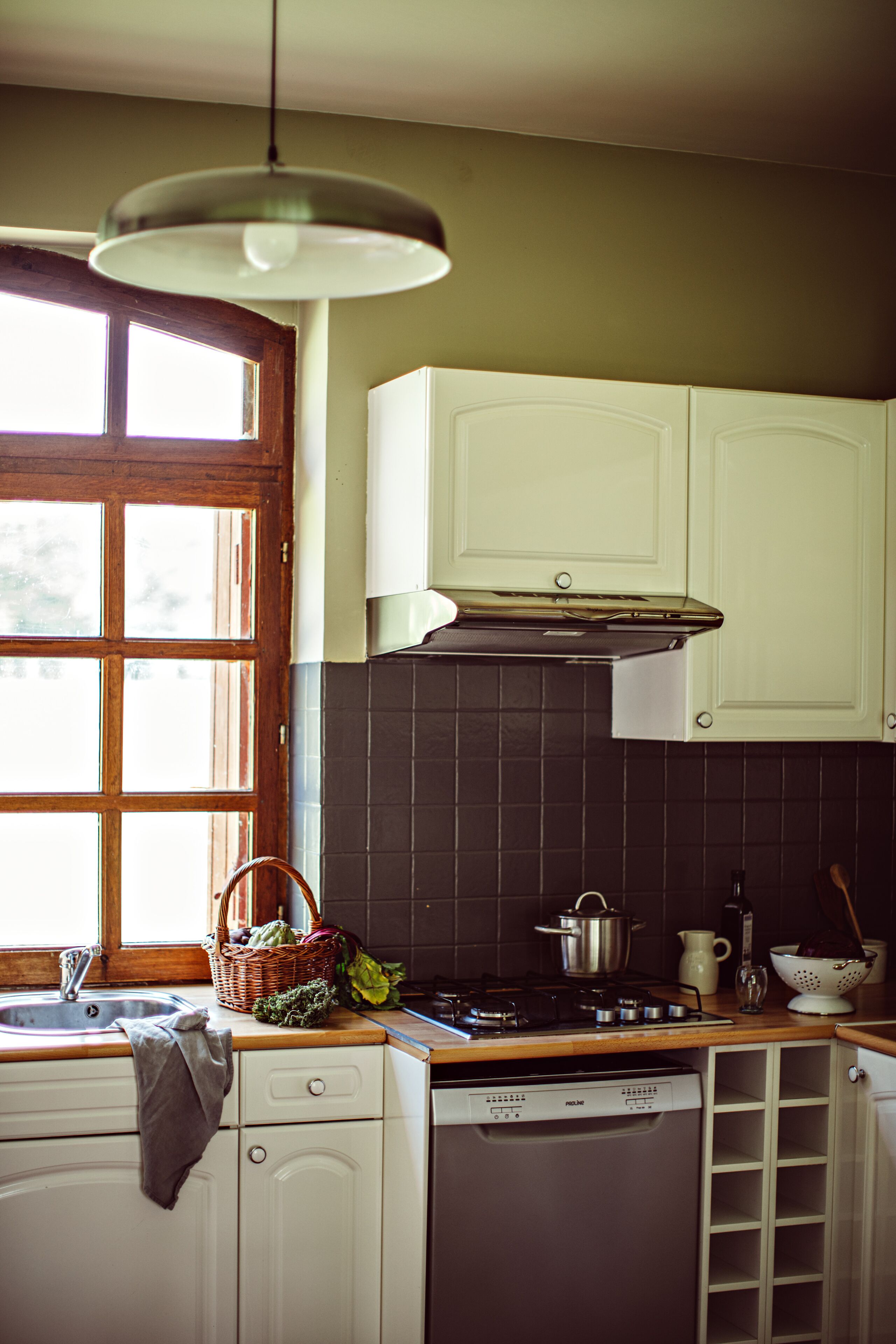 Uma cozinha com armários brancos, painel de azulejos pretos, janela com moldura de madeira, luminária pendente e lava-louças.