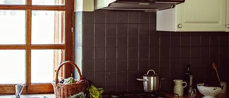 Uma cozinha com armários brancos, painel de azulejos pretos, janela com moldura de madeira, luminária pendente e lava-louças.