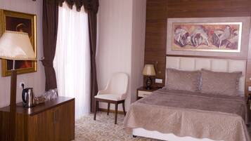 Quarto de hotel com uma cama, uma cadeira, uma escrivaninha com luminária, um quadro na parede e uma janela com cortinas.