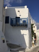 Uma rua estreita com edifícios brancos, uma varanda azul e um pequeno gato no chão.