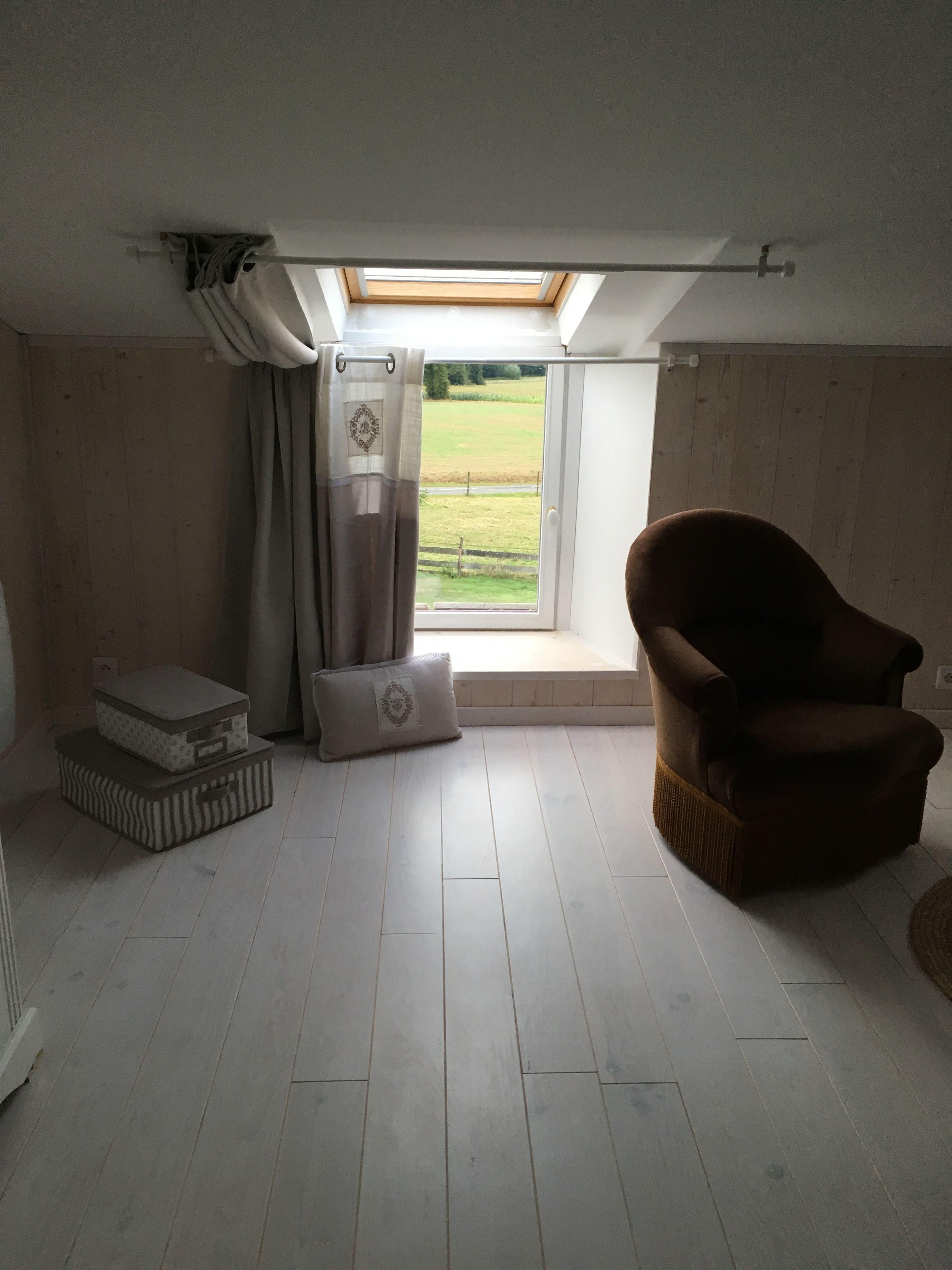 Um quarto com claraboia, uma poltrona marrom, uma mesinha e uma janela com cortinas.