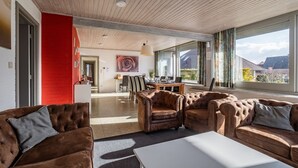 Uma sala de estar espaçosa com teto de madeira, grandes janelas com vista para o exterior e uma área de jantar ao fundo.