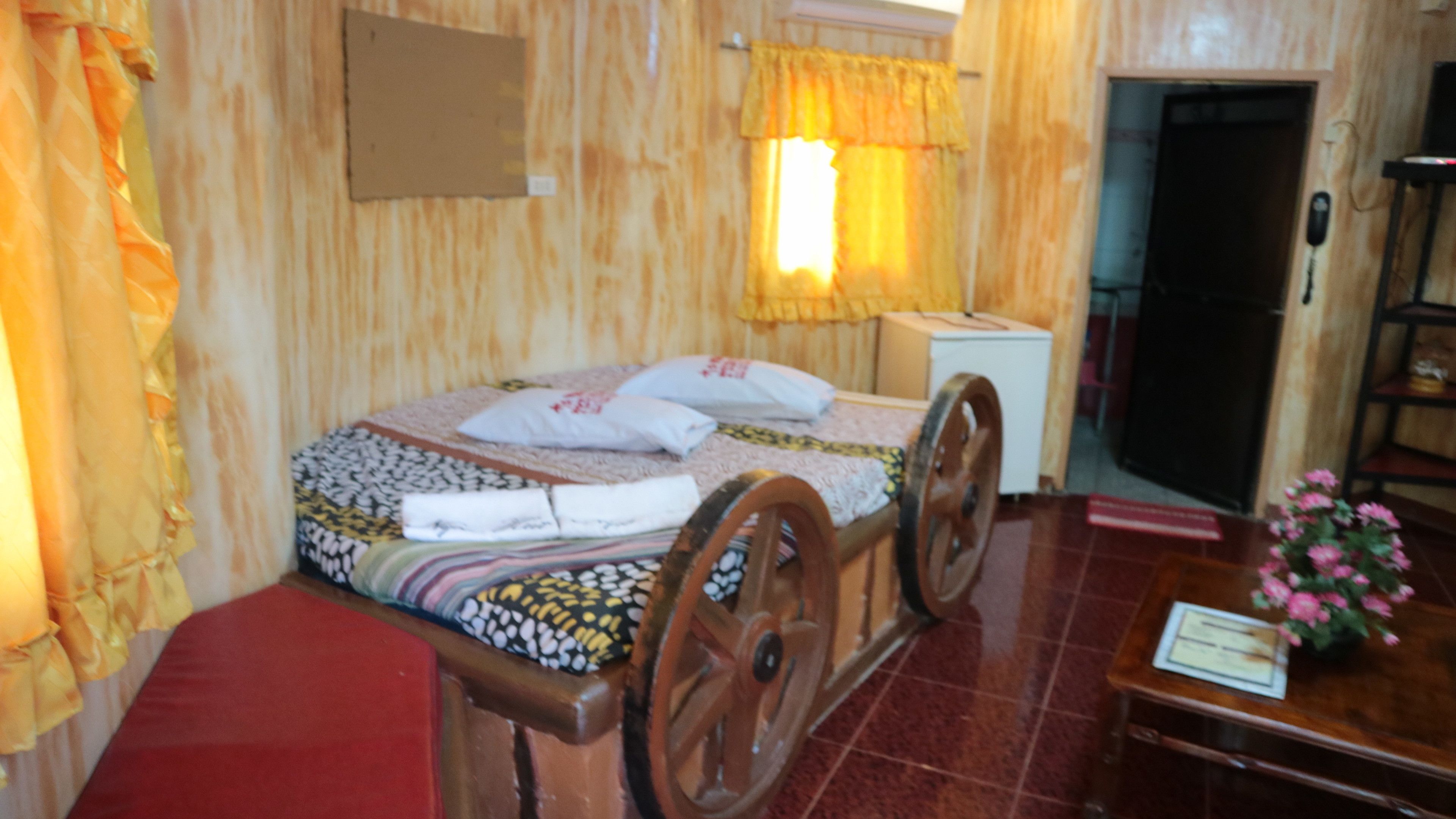 Quarto com cama de madeira, uma pequena mesa de cabeceira, um banco vermelho, guarda-roupa de madeira e uma porta.