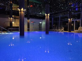 Piscina interna com teto estrelado, espreguiçadeiras e um guarda-sol vermelho.
