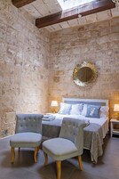 Quarto com paredes de pedra, uma cama, duas poltronas estofadas e um espelho decorativo.