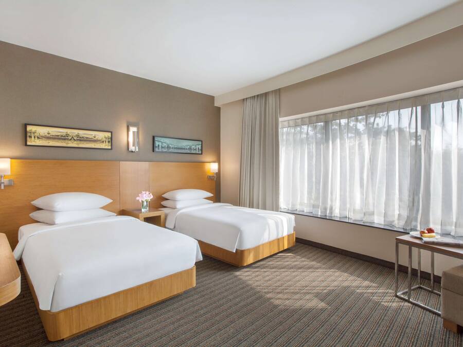Quarto de hotel com duas camas, uma mesa pequena, uma mesa de cabeceira e uma janela com cortinas leves.
