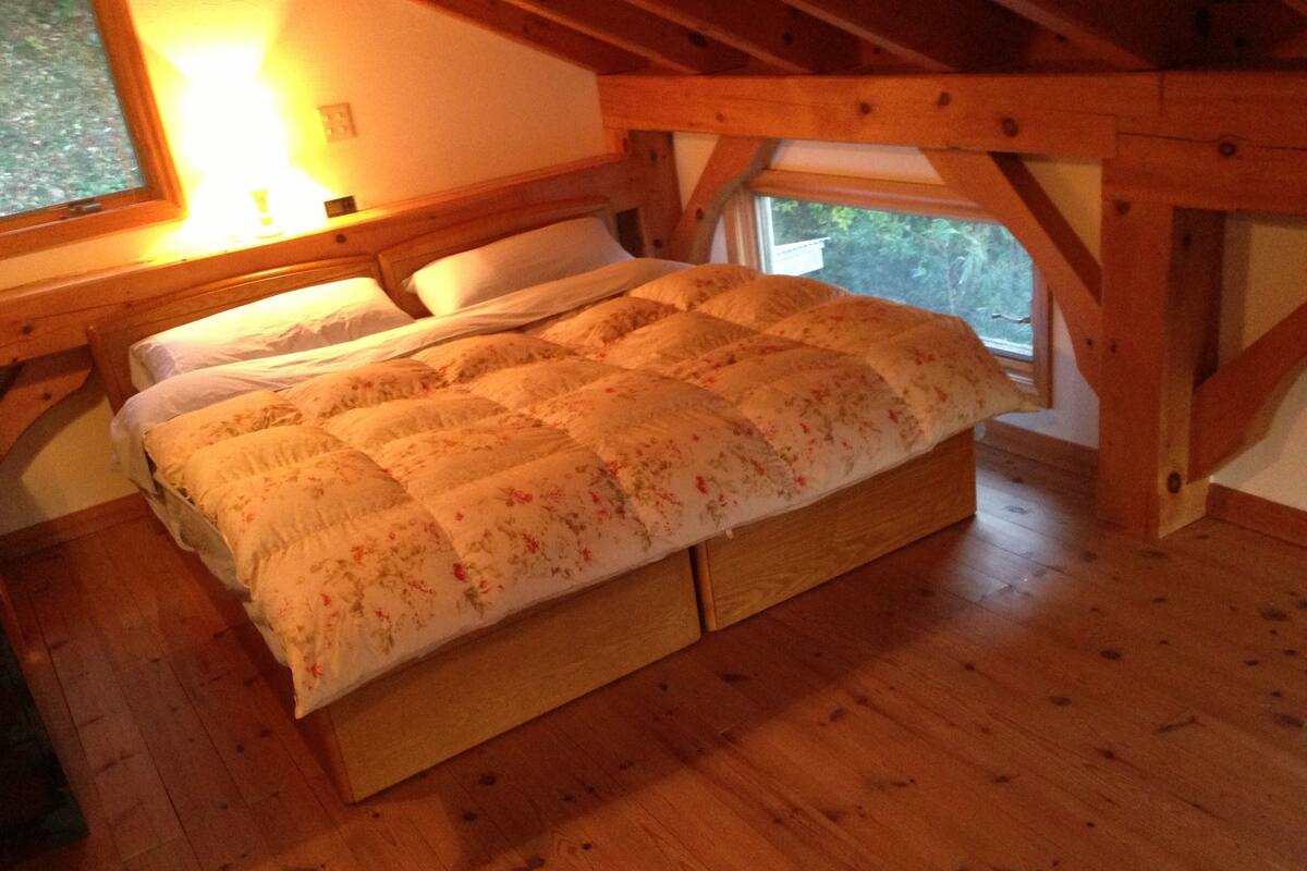 Uma cama de madeira com colcha florida em quarto com piso de madeira e janela.
