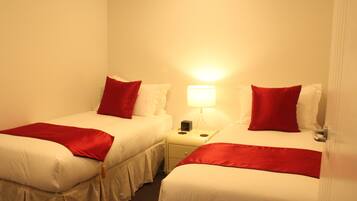 Quarto de hotel com duas camas, cada uma com roupa de cama branca e almofadas vermelhas. Há uma mesa de cabeceira com abajur entre as camas.