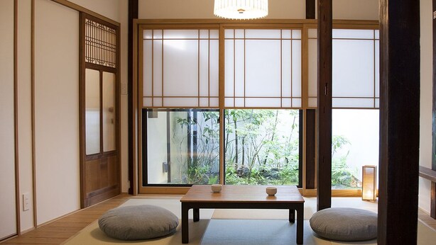 Um quarto japonês tradicional com tatames, uma mesa baixa com duas tigelas e uma janela grande com vista para o verde.