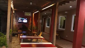 Uma área de estar interna com mesas e cadeiras, uma televisão fixada na parede e uma estrutura de teto em madeira.