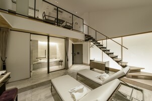 Quarto de hotel moderno com área de estar ampla, uma grande janela e escada que leva a um andar superior.