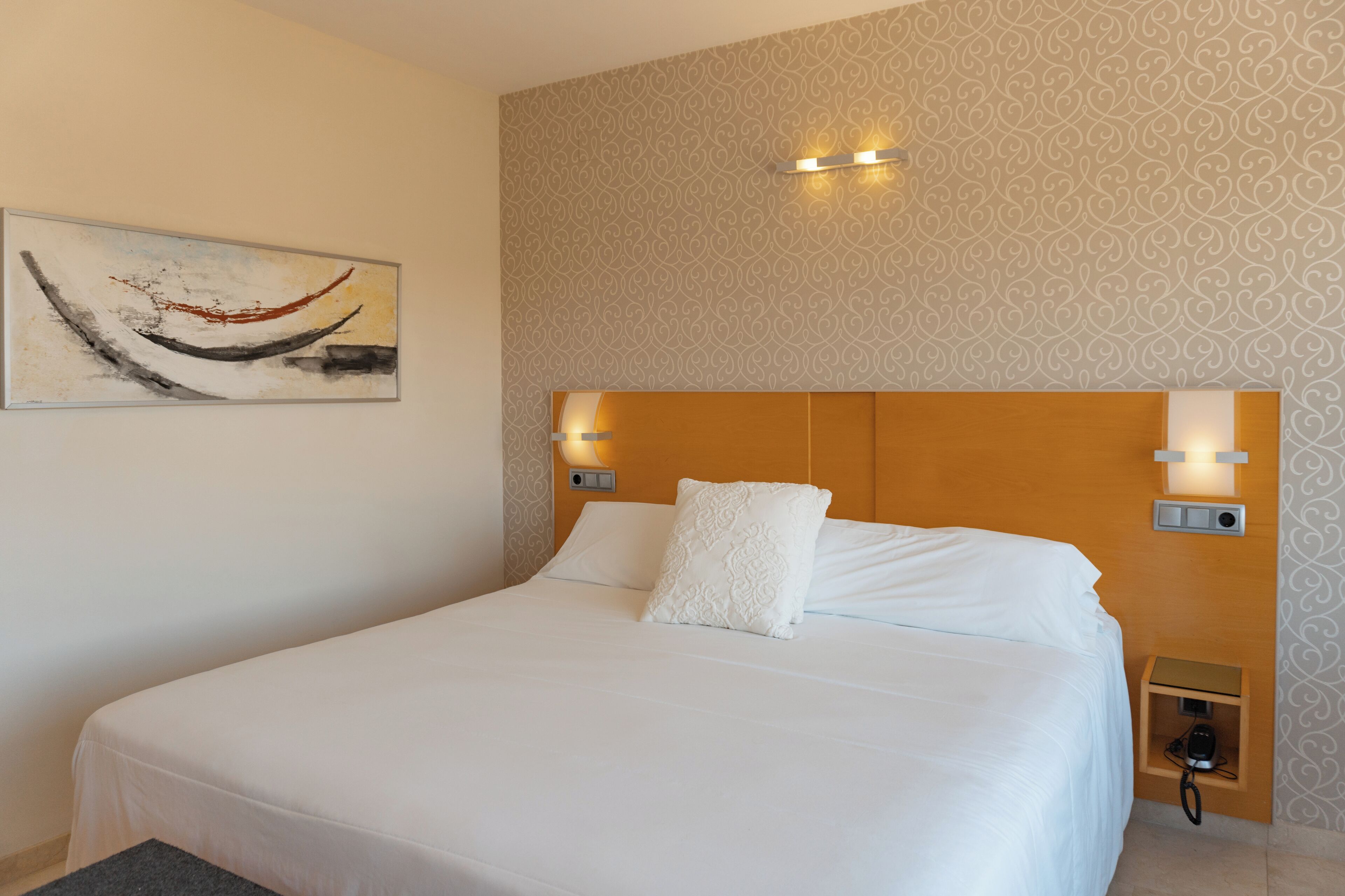 Quarto de hotel com uma cama grande, cabeceira com iluminação integrada, mesa de cabeceira e uma obra de arte pendurada na parede.