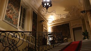 Uma escadaria imponente com degraus cobertos por um tapete vermelho e corrimãos ricamente decorados, que leva a um teto alto com molduras ornamentais e um lustre.