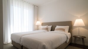 Quarto de hotel com duas camas, uma janela grande com cortinas finas e luminárias laterais.