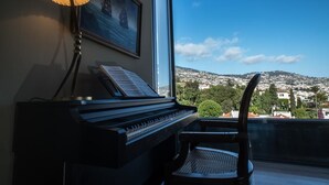 Um piano com partituras, uma cadeira, um abajur e um quadro em uma varanda com vista para uma paisagem urbana e colinas.