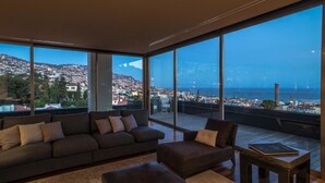 Sala moderna com um sofá grande, uma mesa de centro e vista para a cidade e o mar ao entardecer.
