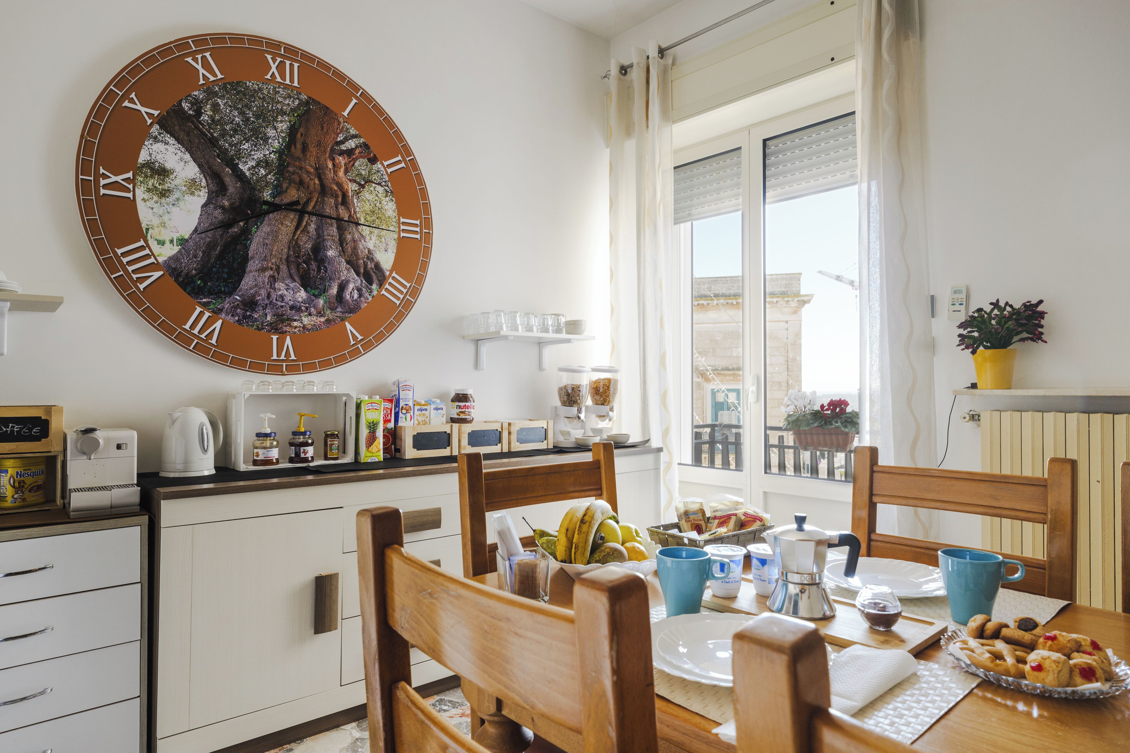 Uma cozinha com um relógio de parede grande, cadeiras de madeira, uma mesa de jantar com itens de café da manhã e uma janela com vista.