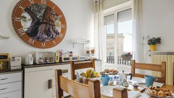 Uma cozinha com um relógio de parede grande, cadeiras de madeira, uma mesa de jantar com itens de café da manhã e uma janela com vista.