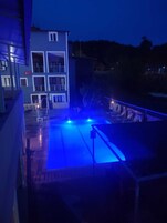 Uma piscina com iluminação azul, um edifício com varandas e vista para árvores.