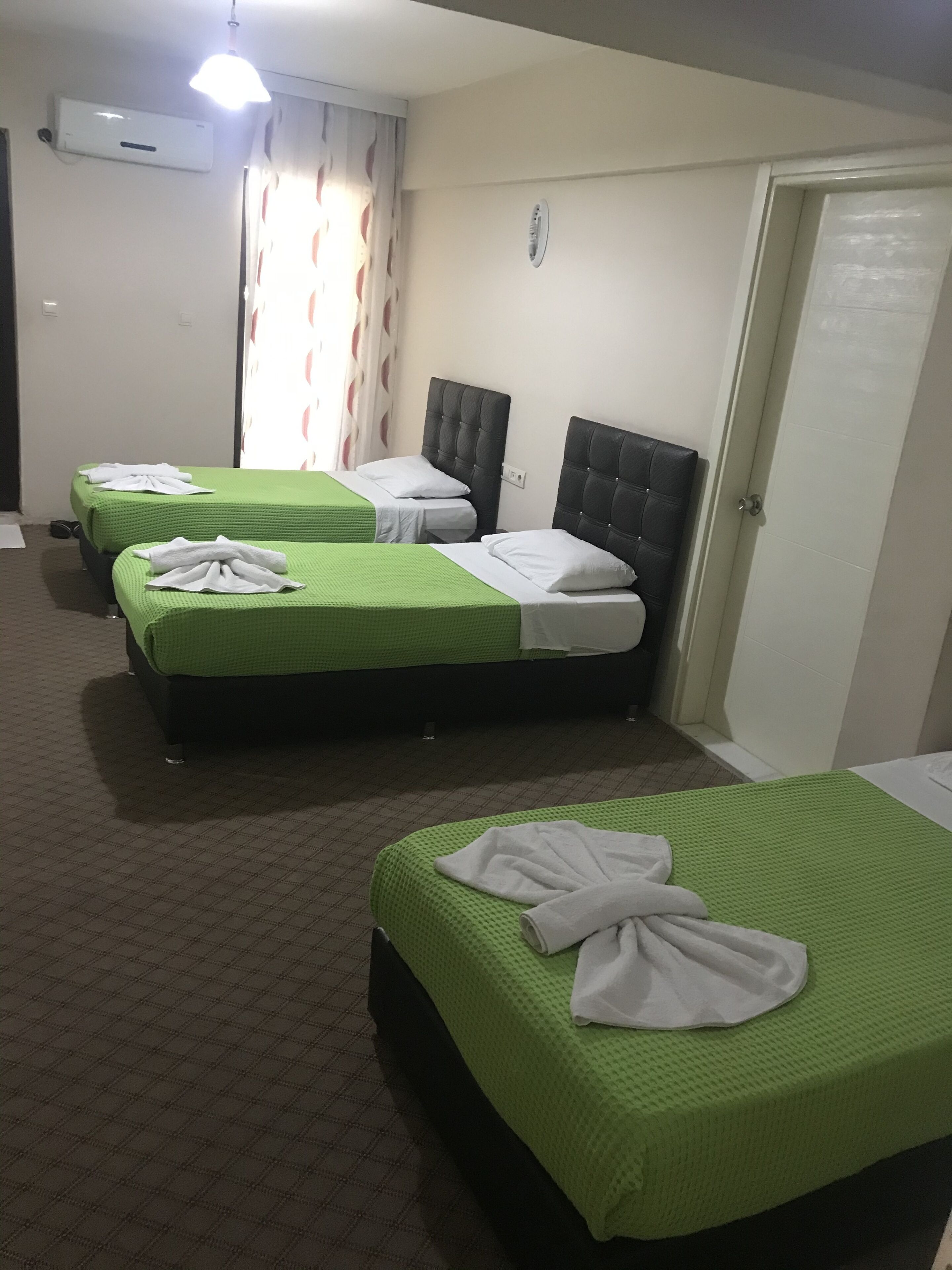 Quarto de hotel com três camas, cada uma com roupa de cama verde e almofadas brancas. Há um ventilador de teto, um ar condicionado e uma porta com puxador.