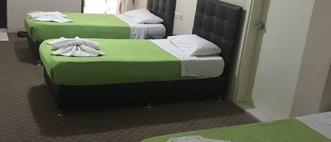 Quarto de hotel com três camas, cada uma com roupa de cama verde e almofadas brancas. Há um ventilador de teto, um ar condicionado e uma porta com puxador.