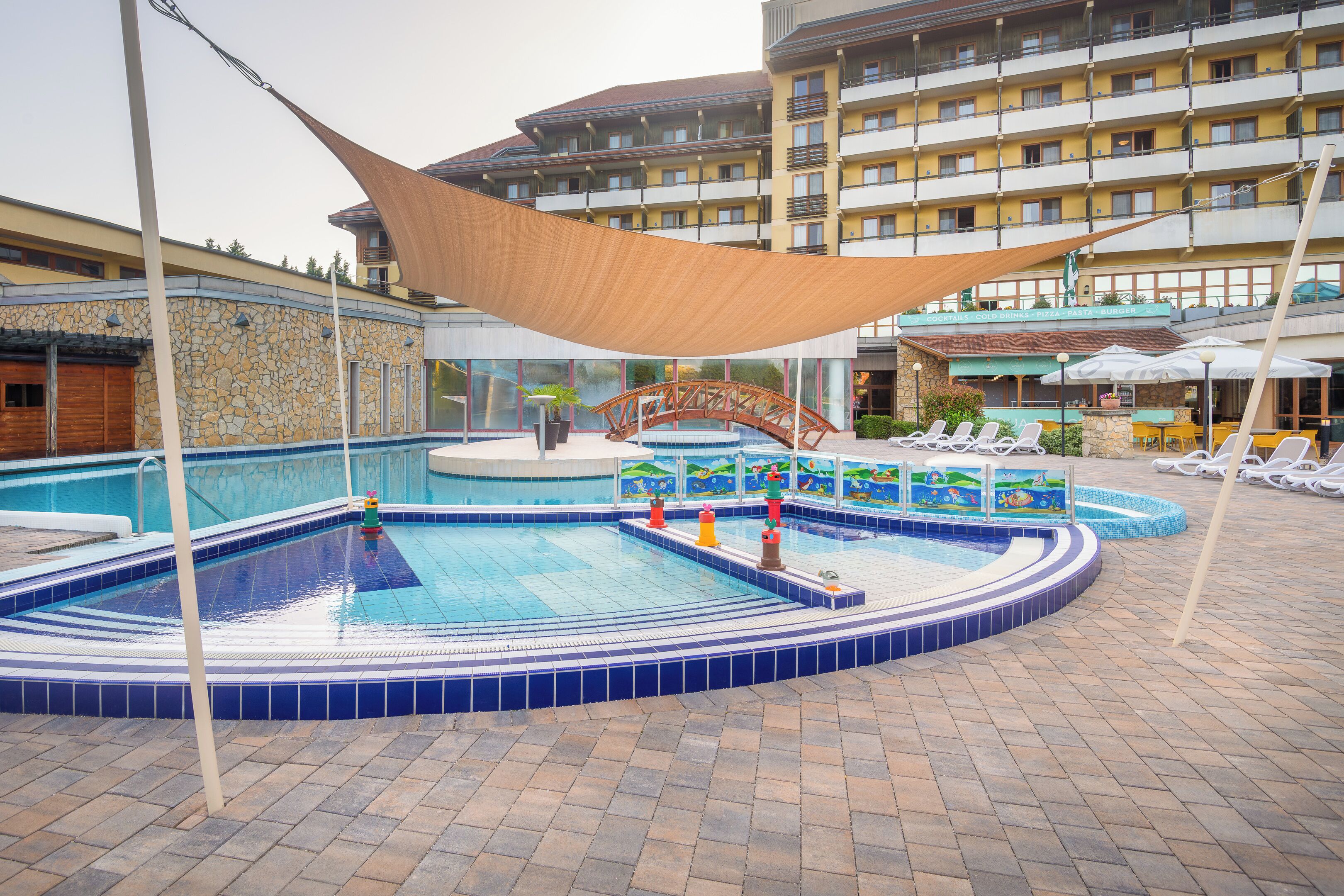 Área de piscina com uma ponte, cadeiras de lounge e um edifício ao fundo.