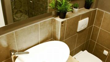 Casa de banho com lavatório branco, sanita e zona de duche com azulejos estampados. Há plantas numa prateleira acima da sanita.