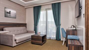 Quarto de hotel moderno com sofá, mesa de centro, escrivaninha com cadeira e uma janela grande com cortinas.