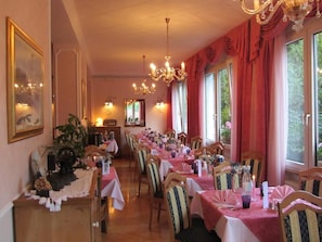 Uma sala de jantar com mesas postas para vários convidados, cadeiras de madeira e um candelabro.