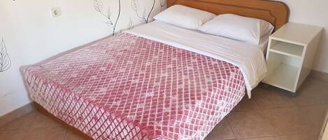 Uma cama com colcha xadrez rosa, cabeceira de madeira, travesseiros brancos e uma mesa de cabeceira com abajur branco.