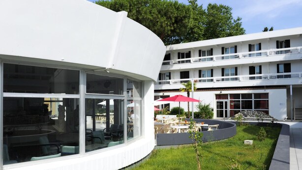 Um edifício moderno com grandes janelas e fachada curva, um pátio com área de estar ao ar livre e uma estrutura de vários andares com varandas ao fundo.