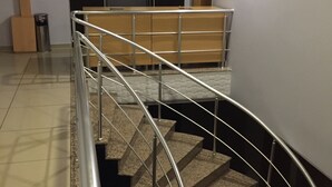 Uma escadaria com guarda-corpos e corrimão de metal, que leva a um patamar com um banco de madeira e uma porta de elevador preta.