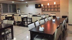 Uma área de refeições com várias mesas e cadeiras, um relógio na parede e uma televisão montada acima de uma estante com objetos decorativos.