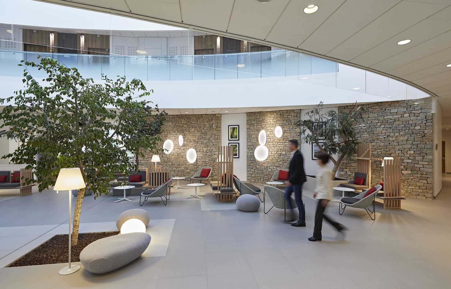 Um lobby moderno e interno com teto de vidro, paredes de pedra e áreas de estar contemporâneas.