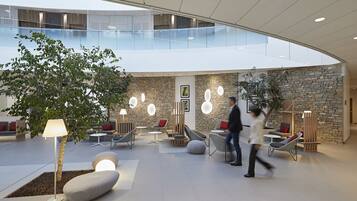 Um lobby moderno e interno com teto de vidro, paredes de pedra e áreas de estar contemporâneas.