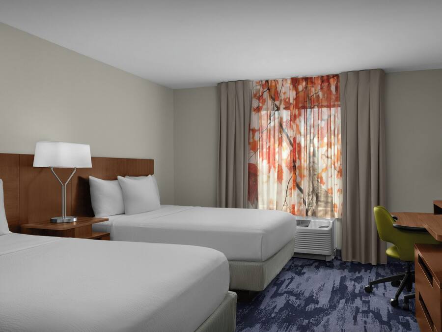 Quarto de hotel com duas camas, uma escrivaninha, uma cadeira, uma televisão e uma janela com cortinas.