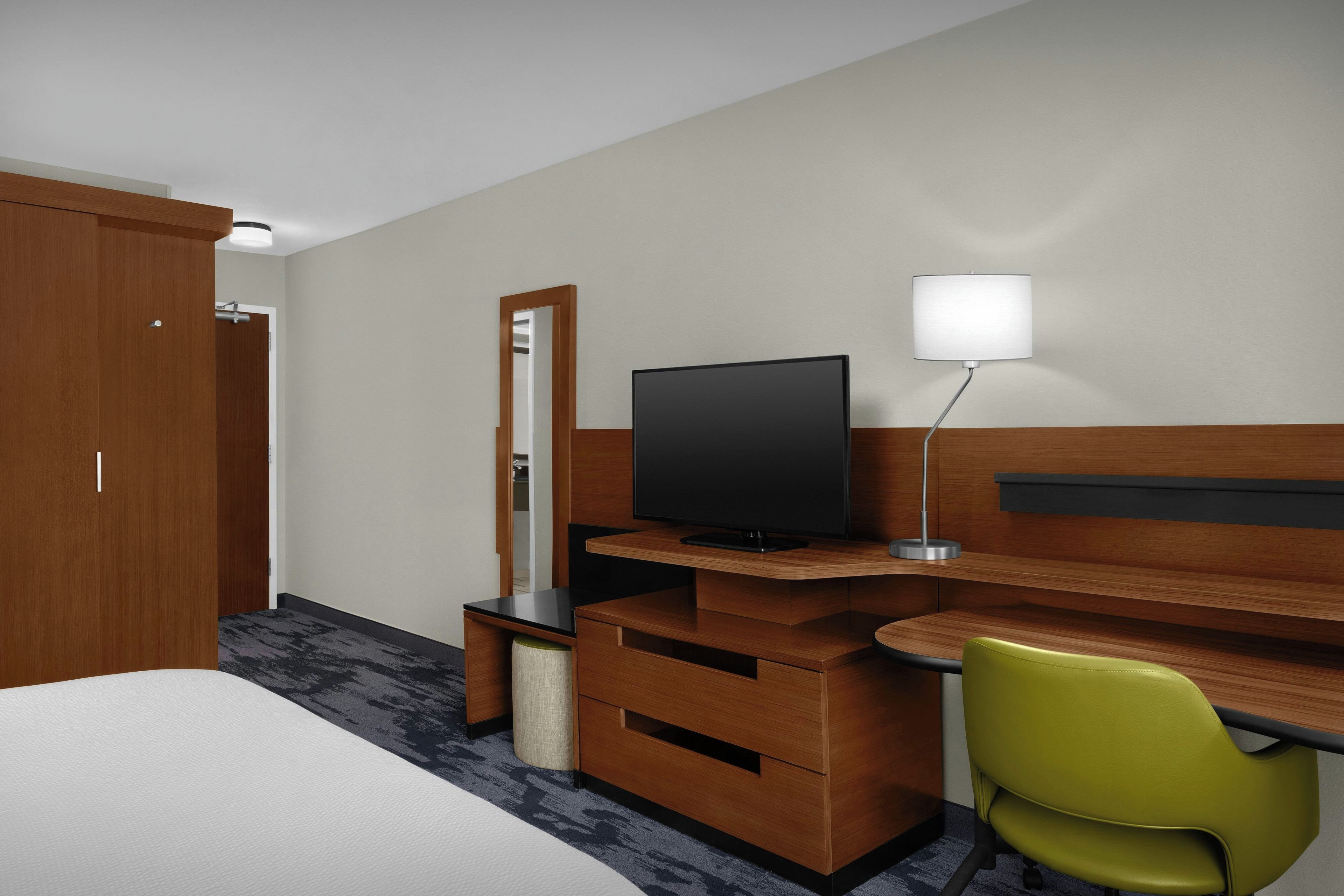 Quarto de hotel com escrivaninha de madeira, televisão, luminária, cadeira verde e armário.