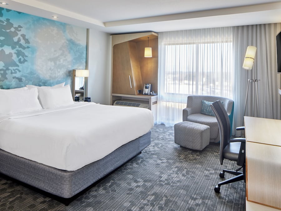 Quarto de hotel com uma cama grande, uma escrivaninha, uma cadeira, uma televisão e uma janela com cortinas.
