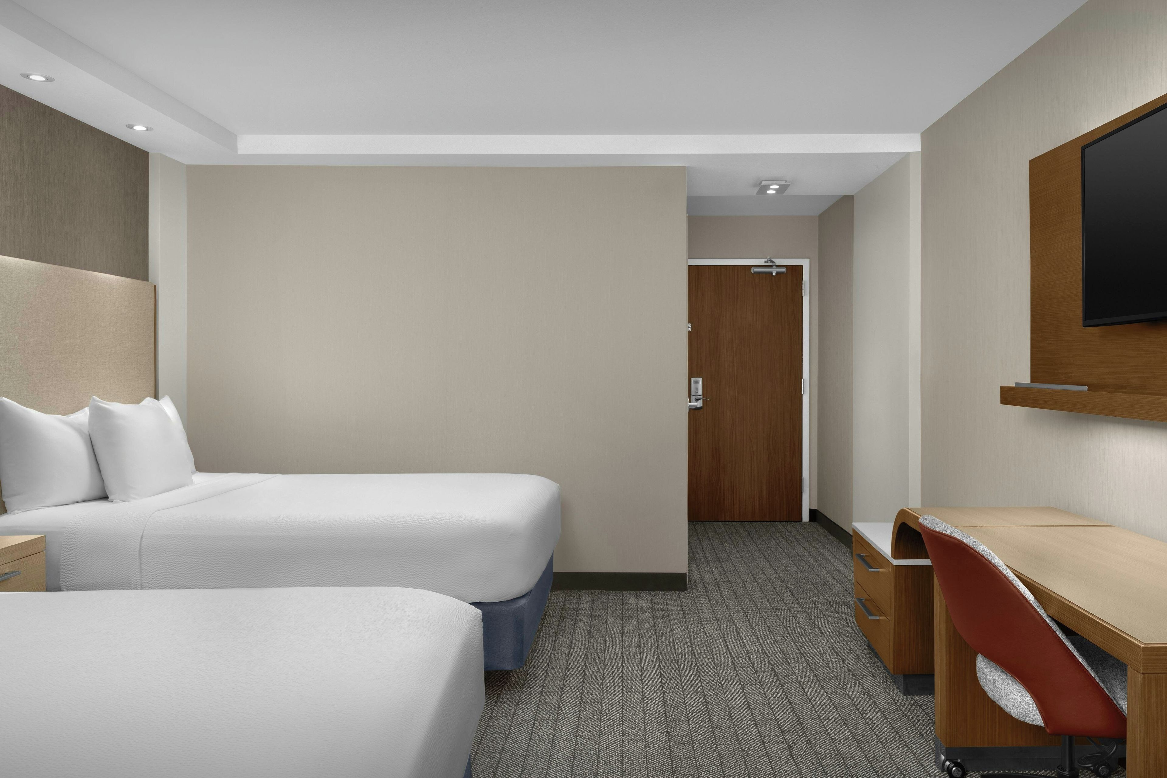 Quarto de hotel com duas camas, uma escrivaninha, uma cadeira, uma televisão e uma porta.
