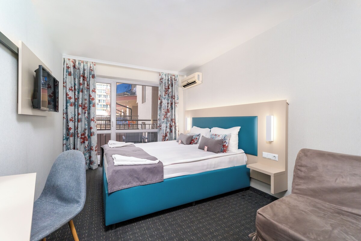 Quarto de hotel com cama, escrivaninha, cadeira e televisão.