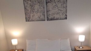 Uma cama bem arrumada com lençóis brancos e um edredom cinza, ladeada por duas mesas de cabeceira com luminárias, em um quarto com paredes lisas e duas obras de arte emolduradas acima da cama.