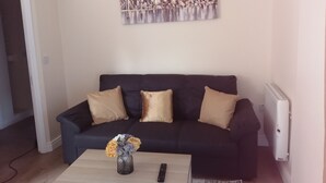 Uma sala de estar com um sofá de cor escura, almofadas bege, um vaso com flores sobre uma mesa de centro de madeira, um controle remoto e um quadro na parede.