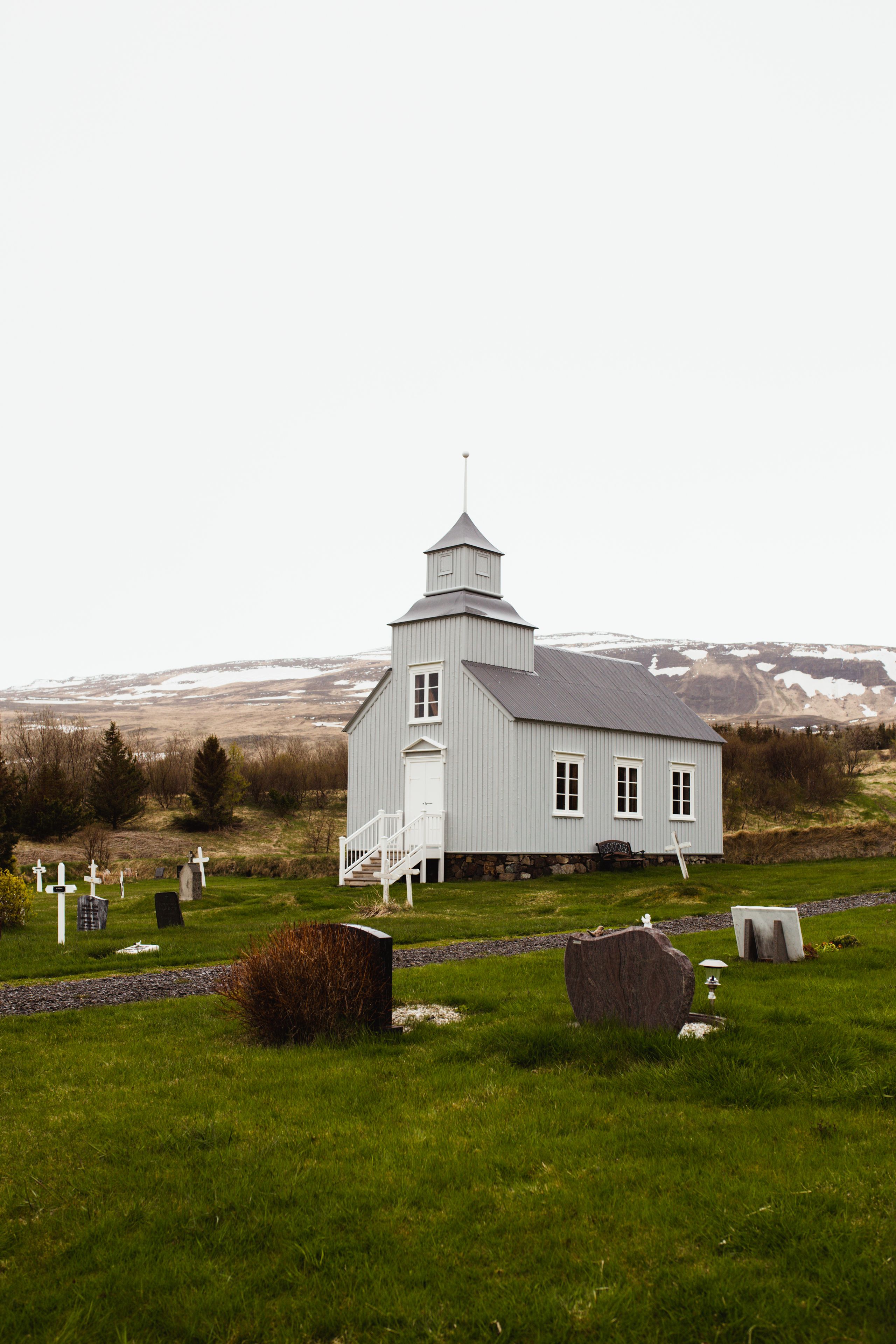 Uma igreja branca com um campanário, rodeada por um campo gramado com lápides e uma montanha ao fundo.