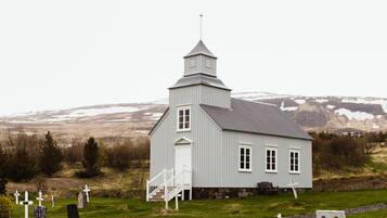 Uma igreja branca com um campanário, rodeada por um campo gramado com lápides e uma montanha ao fundo.